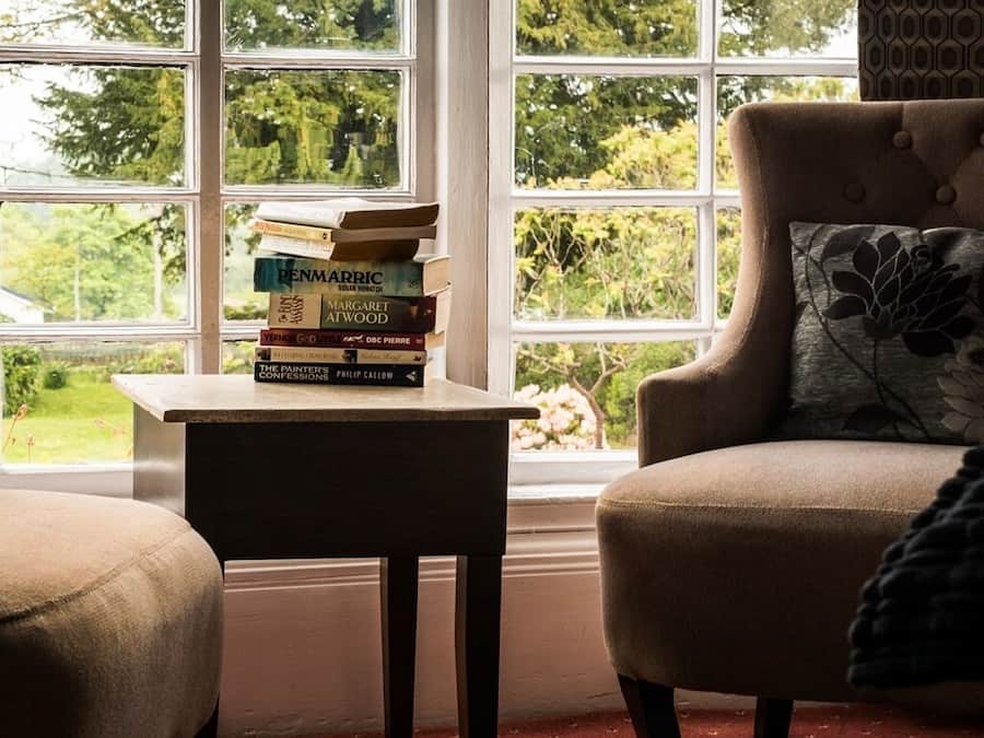 Quarto com janela, pilha de livros sobre uma mesa, cadeira com almofada florida e vista para o verde lá fora.