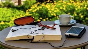 Mesa redonda com livro, óculos, um smartphone e uma xícara, com o cenário de uma praia com guarda-sóis e palmeiras.