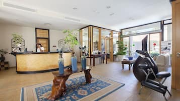 Uma área de recepção com balcão de madeira, uma planta em vaso grande, uma poltrona de massagem e um tapete decorativo.