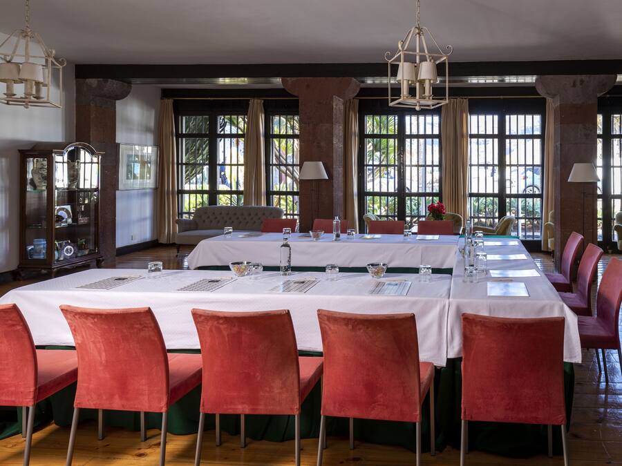 Uma sala de jantar com uma mesa comprida posta para refeição, cercada por cadeiras vermelhas, com janelas amplas e um lustre.