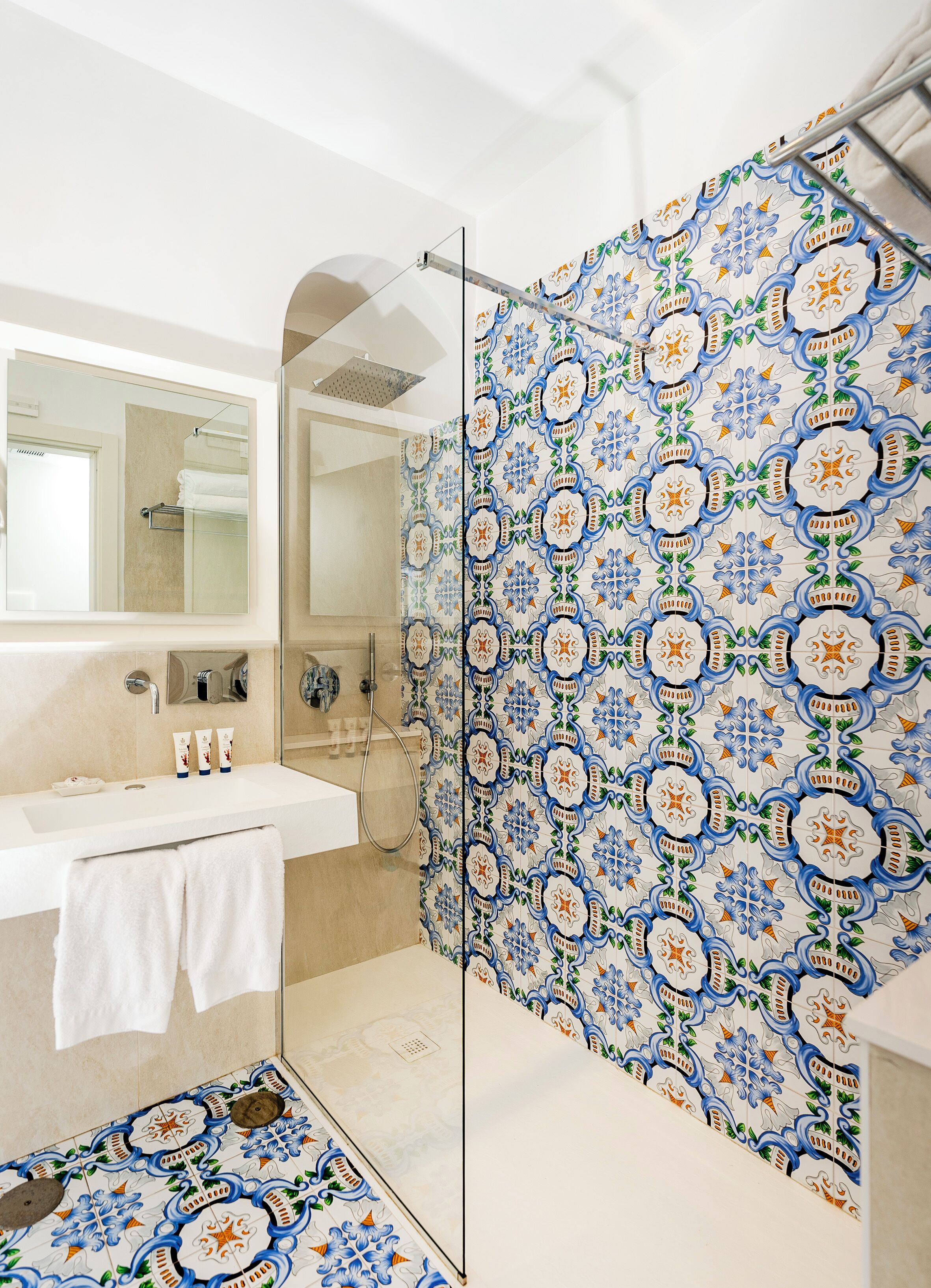 Casa de banho com lavatório branco, espelho e zona de duche com parede em azulejo estampado.