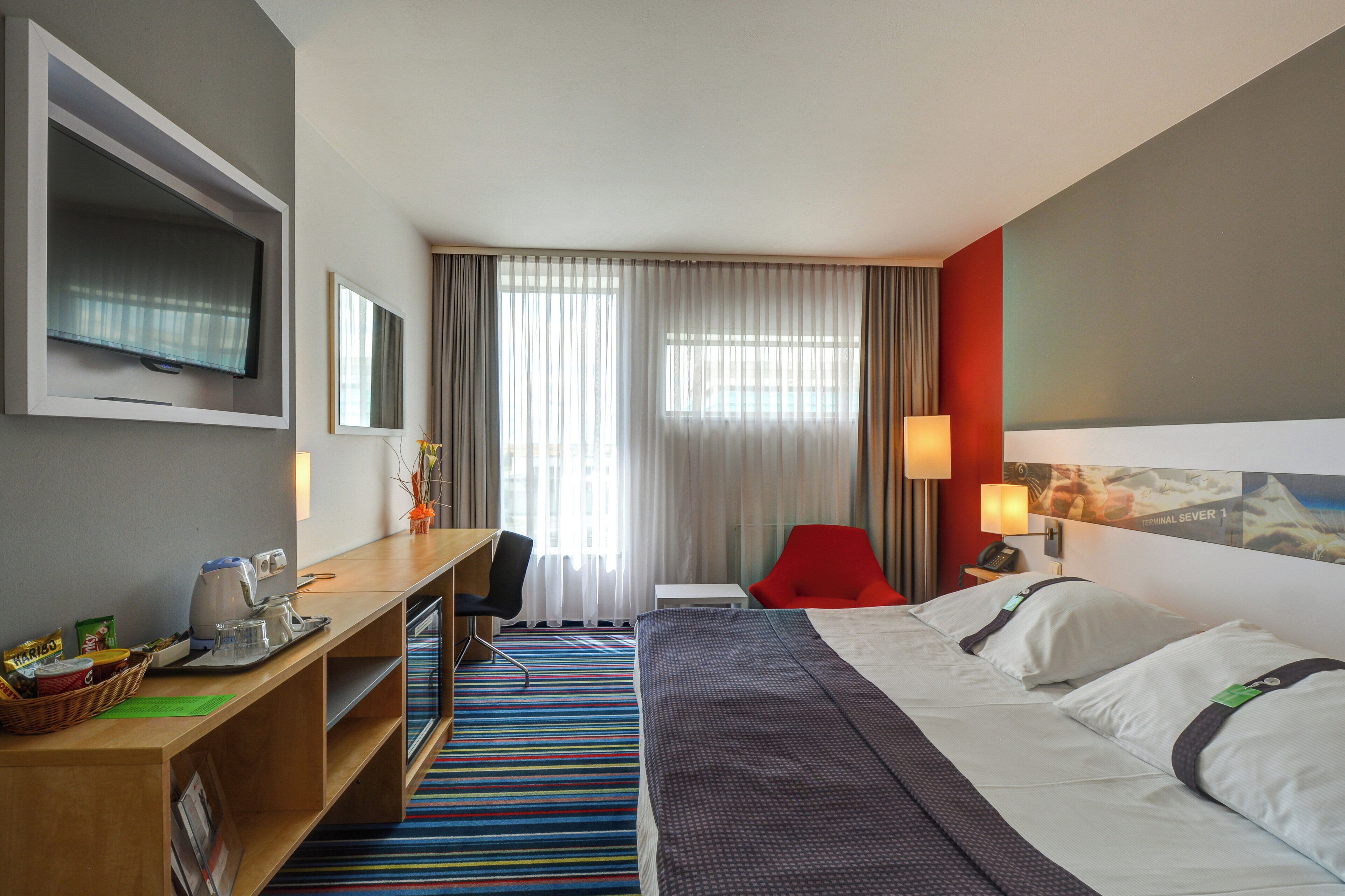 Quarto de hotel com cama, escrivaninha, cadeira, televisão, janela com cortinas e luminária fixada na parede.