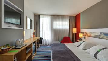 Quarto de hotel com cama, escrivaninha, cadeira, televisão, janela com cortinas e luminária fixada na parede.