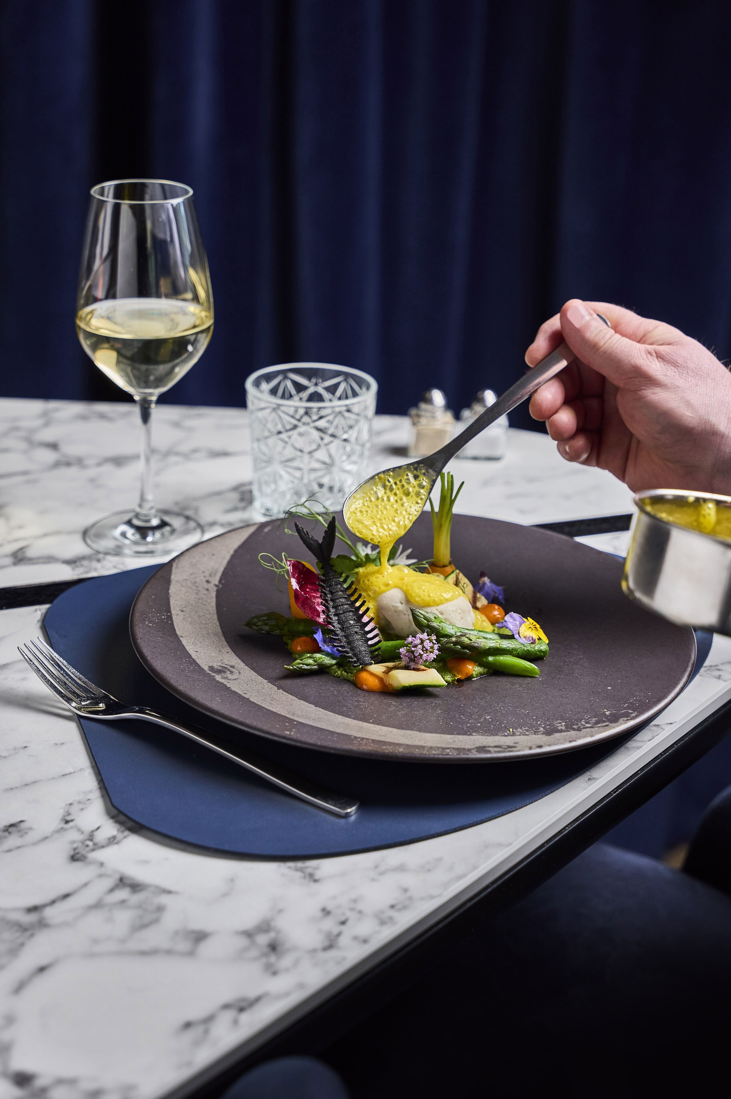 Uma mesa de jantar com um prato de comida, um garfo, um copo de vinho branco e um copo decorativo.