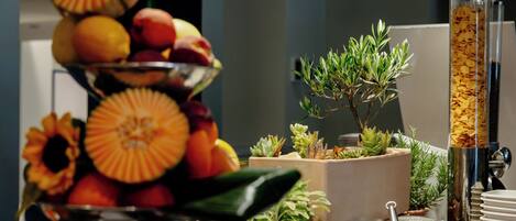 Disposição de buffet com uma exposição de frutas, dispensador de café e pequenas plantas em vasos.