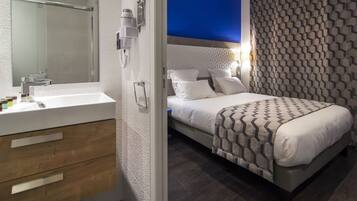 Quarto de hotel moderno com uma cama grande, banheiro com pia branca e toucador de madeira, e parede estampada.