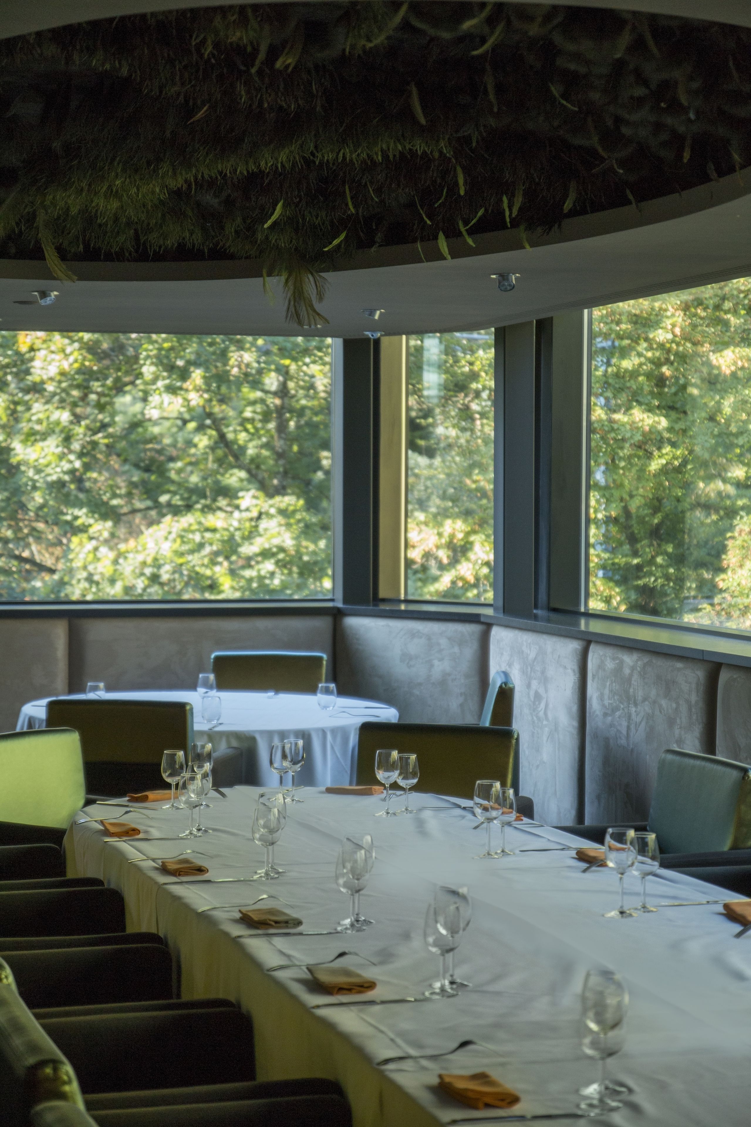 Restaurante com uma mesa comprida posta para refeições, grandes janelas e vista para árvores lá fora.