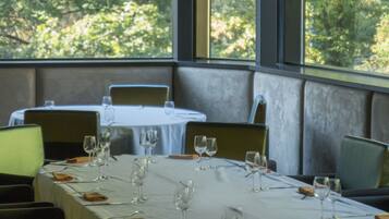 Restaurante com uma mesa comprida posta para refeições, grandes janelas e vista para árvores lá fora.