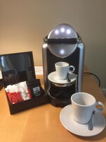 Uma máquina de café com xícara e pires, um suporte com cápsulas de café e uma colher.