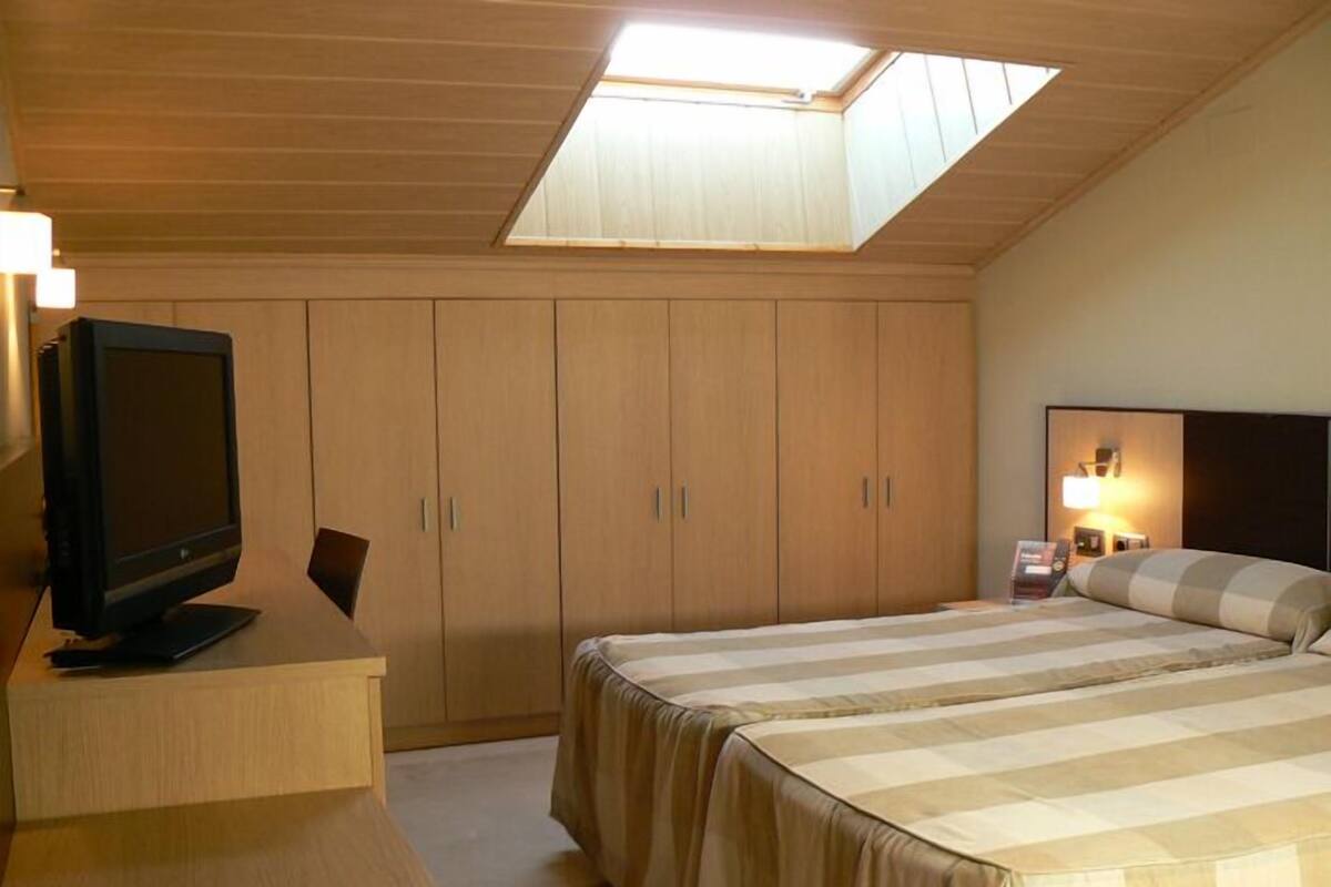Quarto com claraboia, cama, escrivaninha com computador e mesa de cabeceira com abajur.