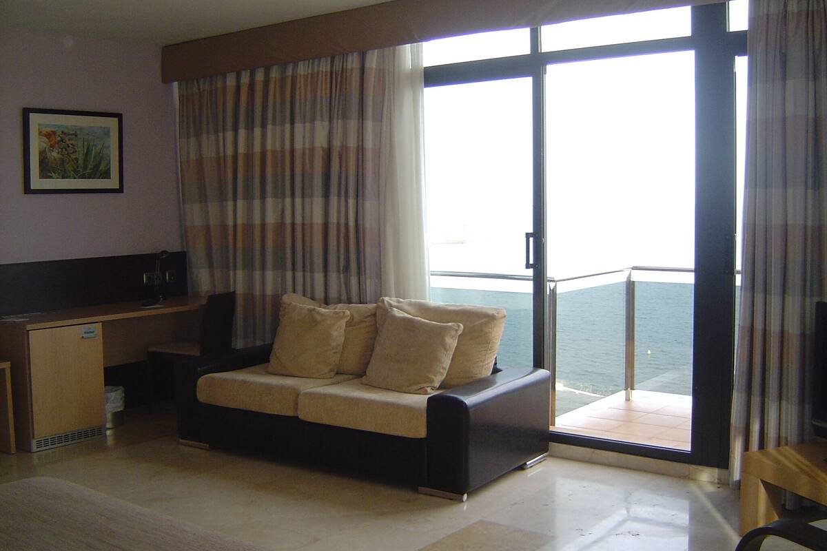 Uma sala de estar com sofá, escrivaninha e vista para o mar através de uma grande janela.
