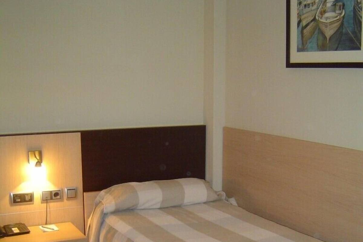 Quarto de hotel com uma cama, uma mesa de cabeceira com telefone e abajur, e um quadro na parede.