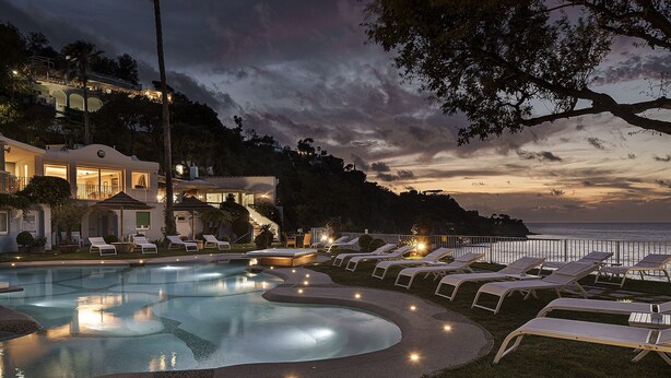 Uma piscina com espreguiçadeiras, um prédio com várias varandas e vista para o oceano ao entardecer.
