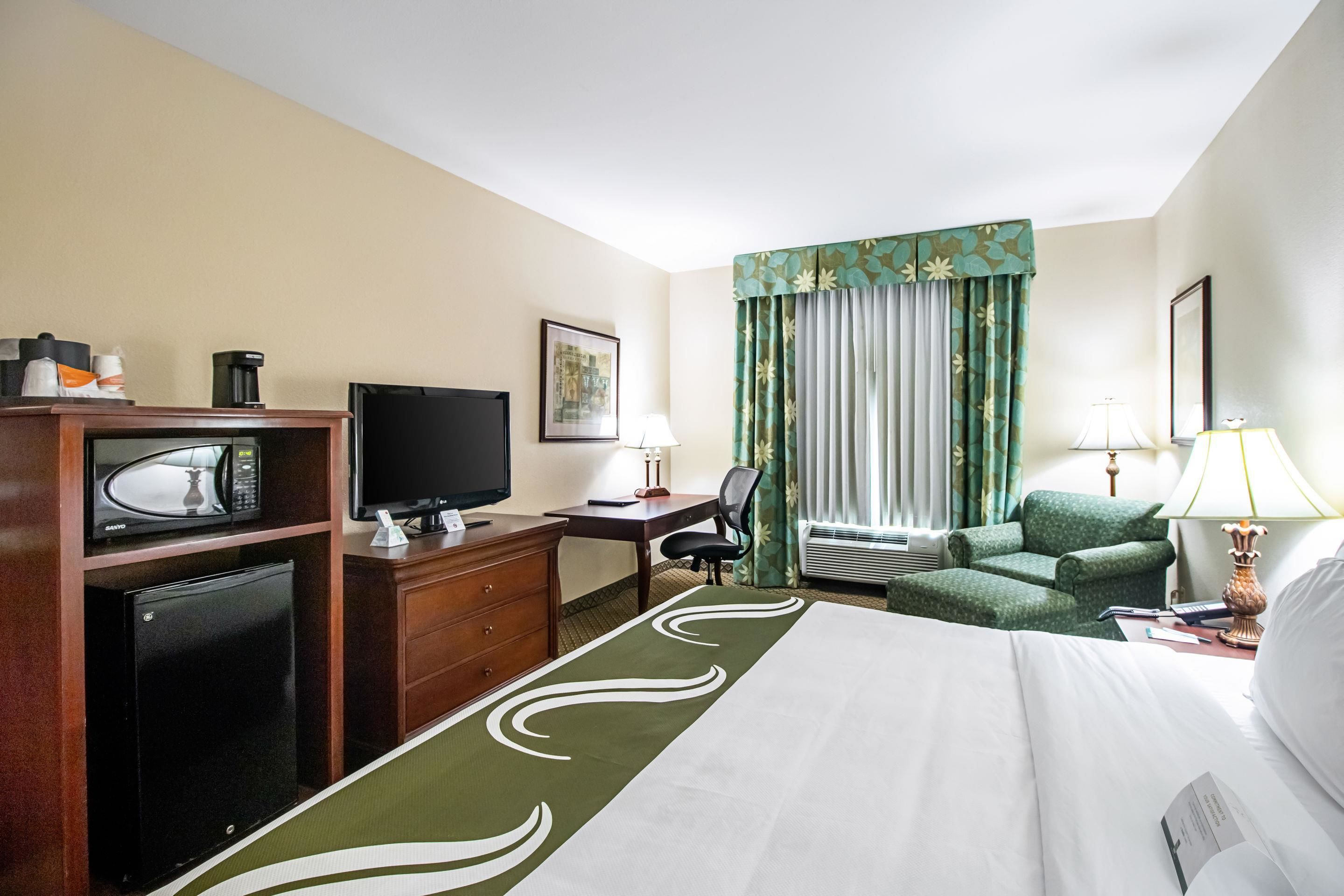 Quarto de hotel com cama, escrivaninha, cadeira, televisão, frigorífico e janela com cortinas.