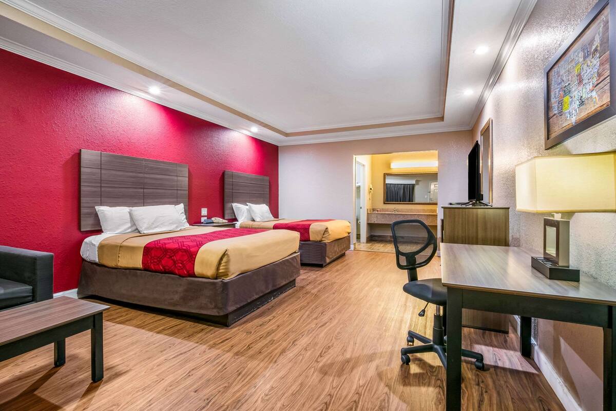 Quarto de hotel com duas camas, uma escrivaninha, uma cadeira e uma mesinha.