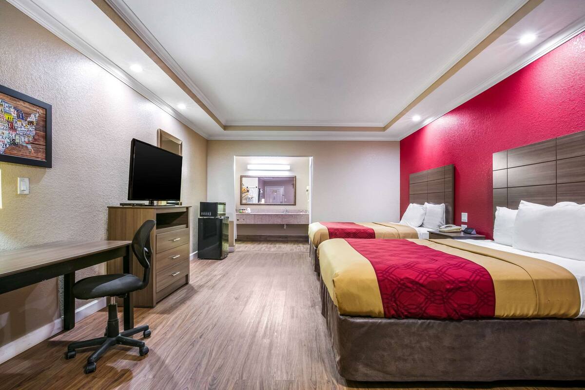 Quarto de hotel com duas camas, uma mesa, uma cadeira, uma televisão e um banheiro visível por uma porta aberta.