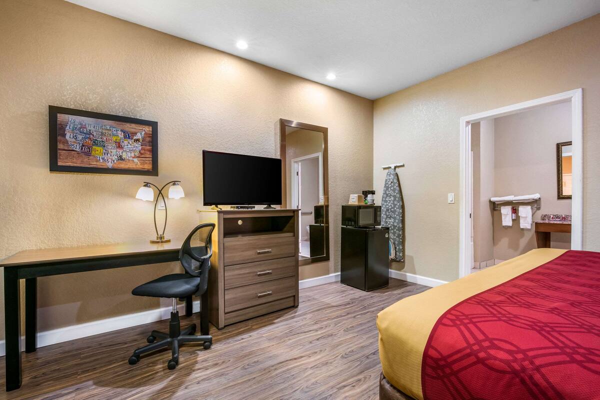 Quarto de hotel com escrivaninha de madeira, televisão, cómoda e cama com colcha vermelha e amarela, além de um quadro pendurado na parede retratando os Estados Unidos.