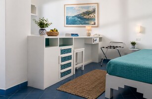 Um quarto com cama, escrivaninha com luminária, cadeira, mesinha com planta e um quadro na parede.