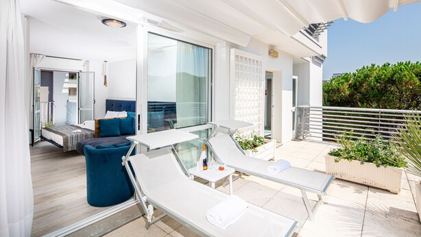 Uma varanda moderna com cadeiras de lounge brancas, um banquinho azul e uma mesinha com uma garrafa e copos.