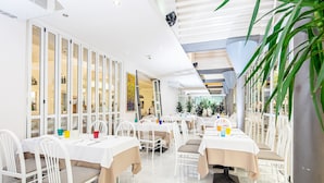 Uma sala de jantar moderna com mesas e cadeiras brancas, grandes janelas e plantas dentro de casa.