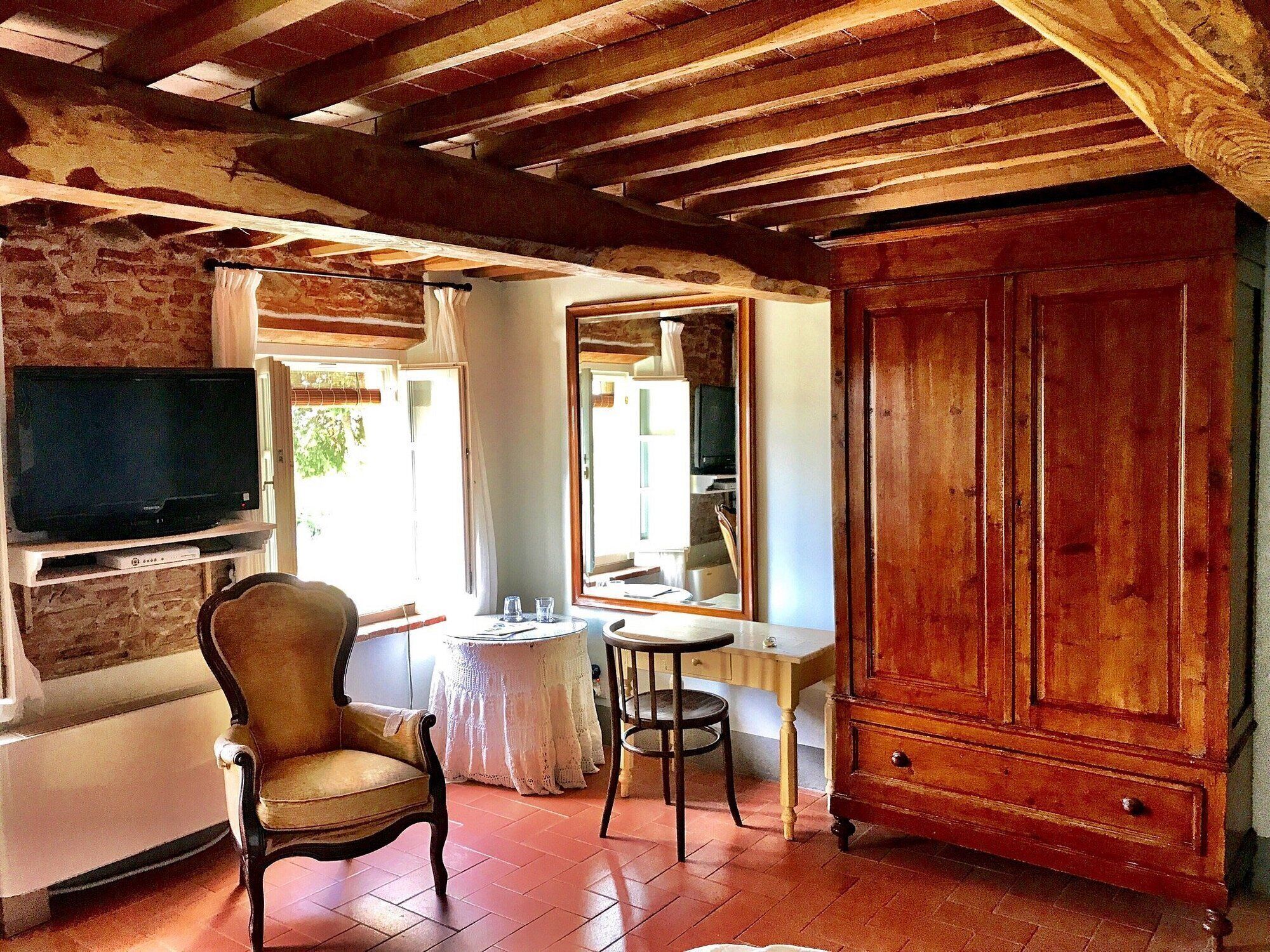 Quarto com televisão em uma estante, um armário de madeira, uma cadeira, uma mesinha e um espelho.