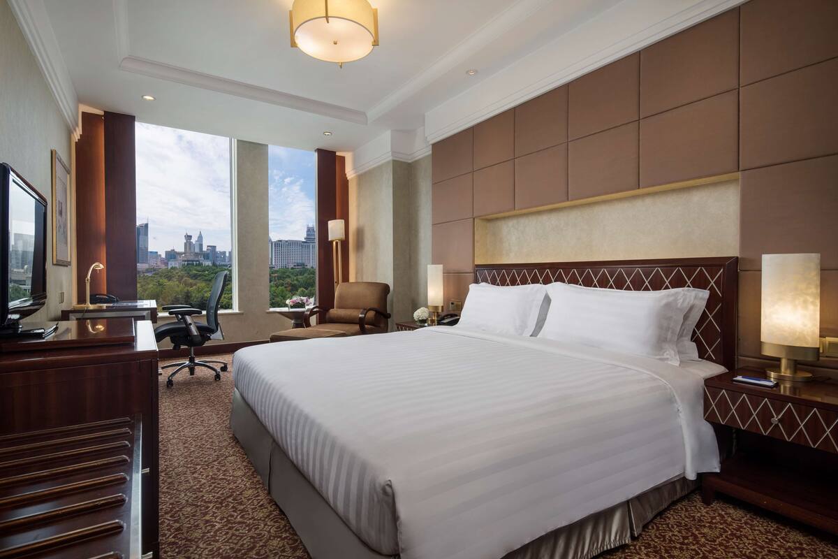Quarto de hotel com uma cama grande, uma mesa com cadeira, televisão e vista para a cidade.