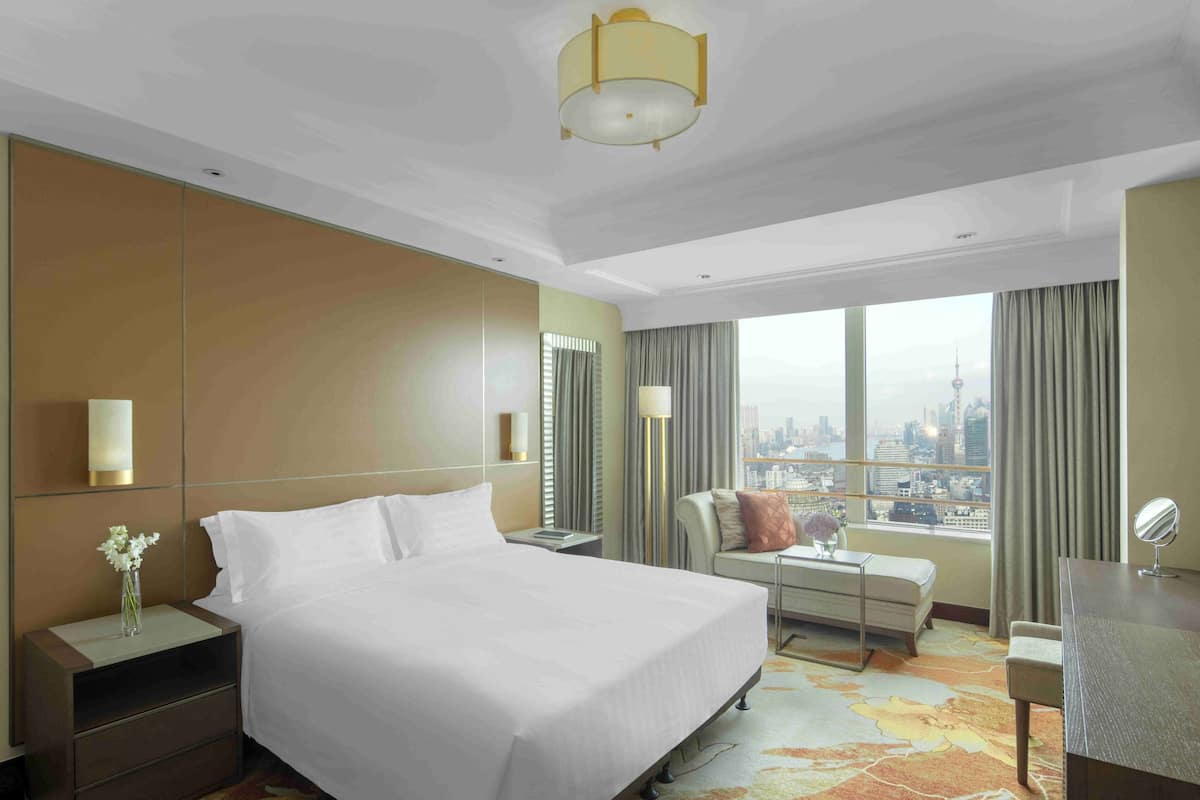 Quarto de hotel com uma cama grande, mesa de cabeceira com um vaso de flores, área de estar com sofá e poltrona, escrivaninha com abajur e vista para a cidade através de grandes janelas.
