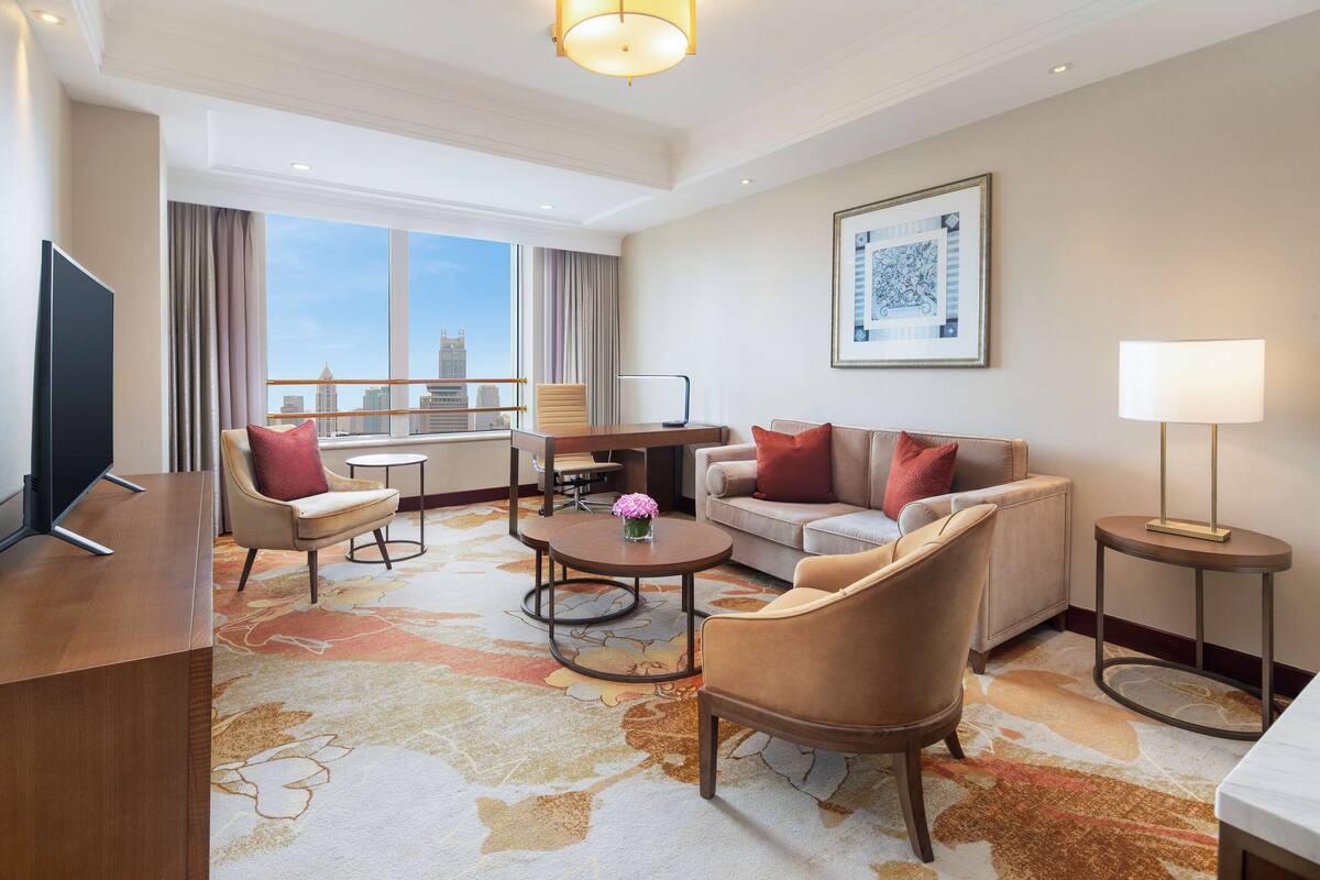 Quarto de hotel com uma janela ampla, televisão de tela plana, sofá, duas poltronas, mesa de centro, mesa lateral com abajur e uma obra de arte emoldurada na parede.
