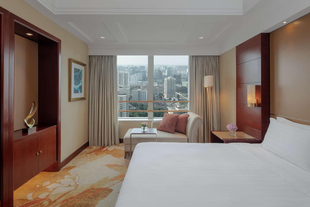 Quarto de hotel com uma cama grande, uma mesa pequena, um sofá, uma mesinha de cabeceira, uma luminária, um quadro na parede e vista da cidade pela janela.