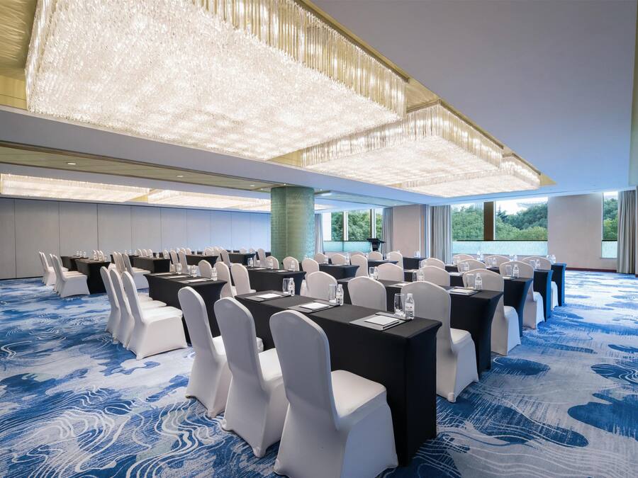 Uma sala de conferências com fileiras de mesas e cadeiras, grandes janelas e um candelabro.