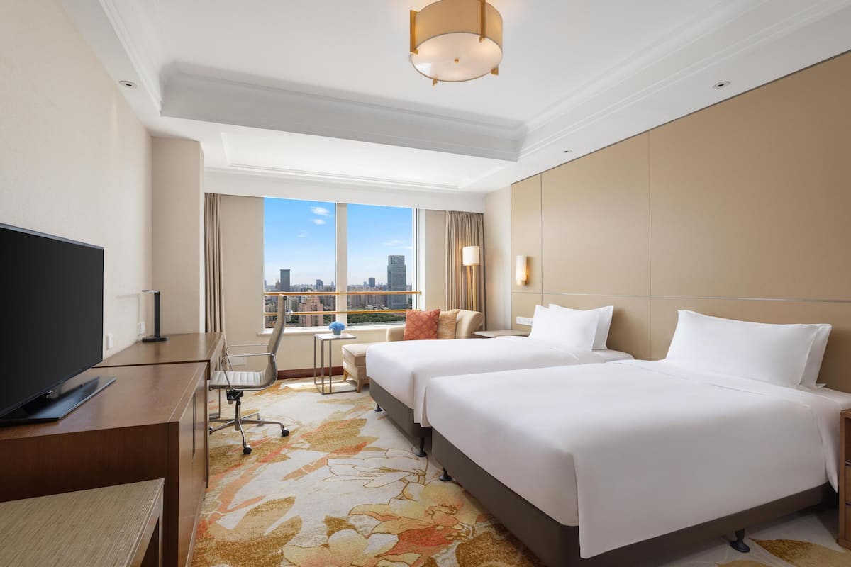 Quarto de hotel com duas camas, uma mesa de trabalho com uma TV, uma mesinha e uma janela grande com vista para a cidade.
