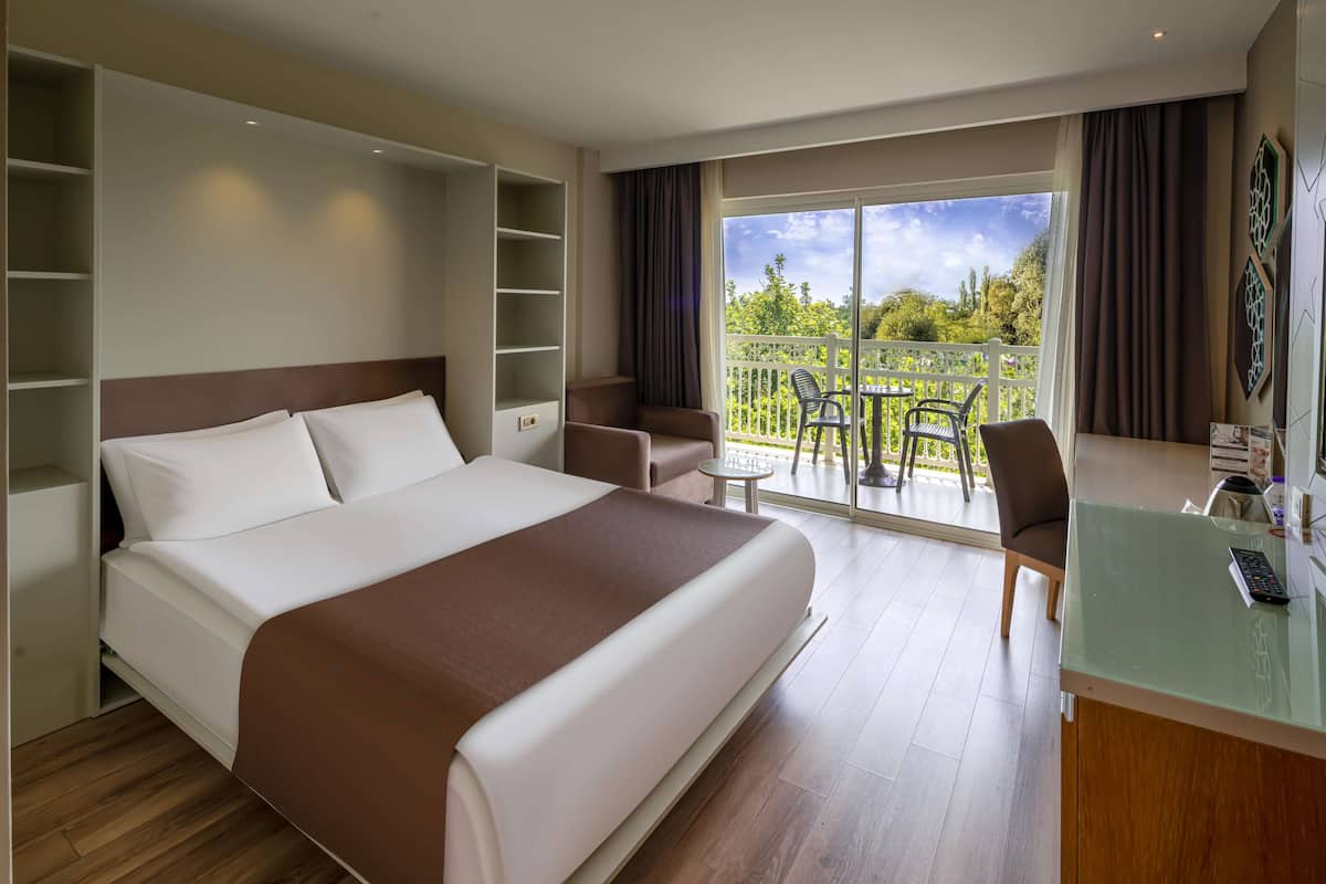 Quarto de hotel com uma cama grande, uma escrivaninha, uma cadeira e varanda com vista para o verde.