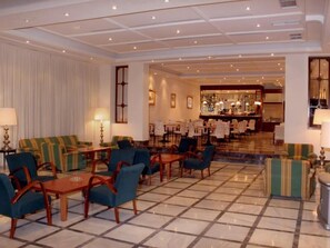 Um hall de hotel com piso xadrez, sofás listrados e uma área de bar ao fundo.