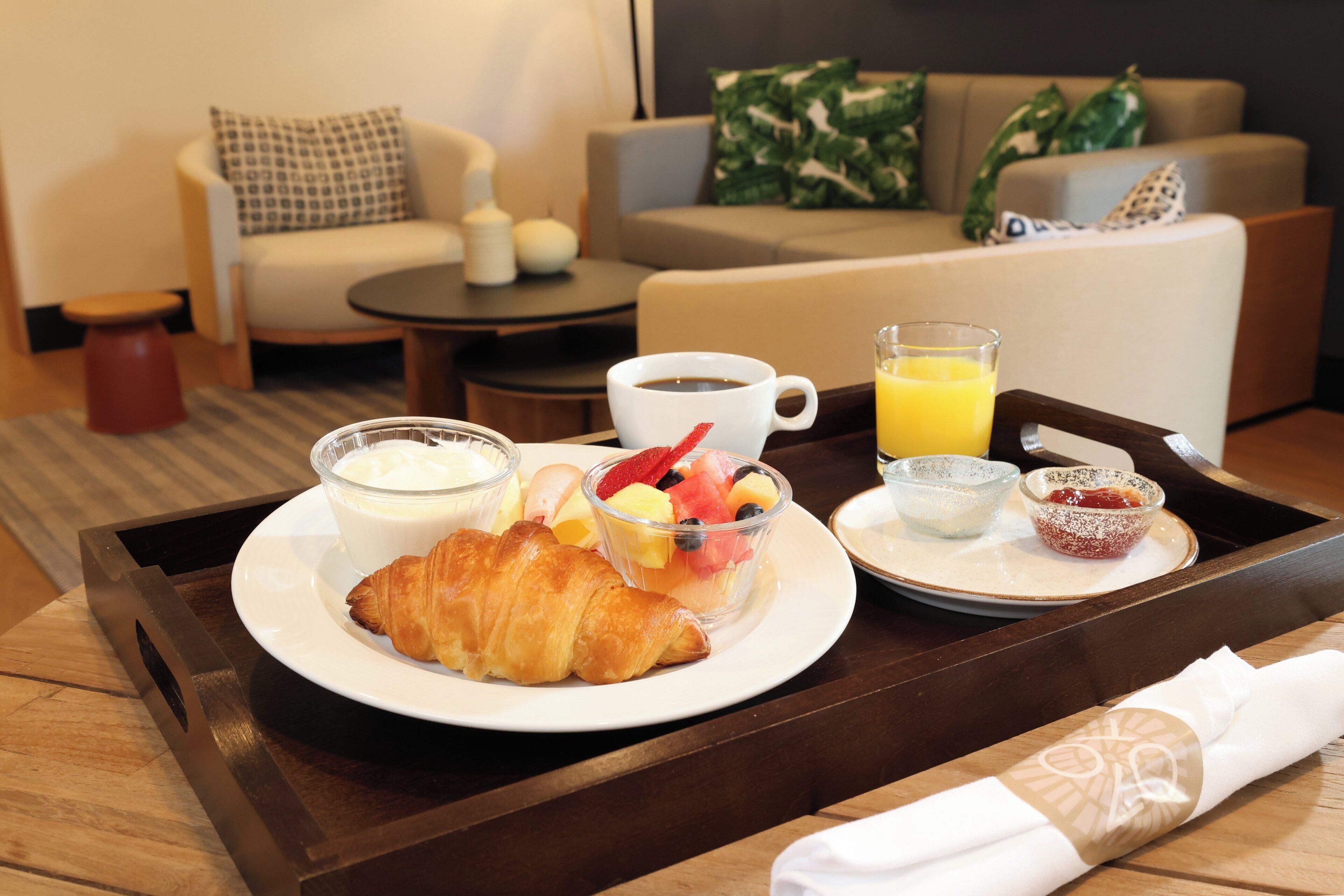 Bandeja de café da manhã com croissant, iogurte, frutas, café e suco em quarto de hotel, com sofá e uma mesa pequena.