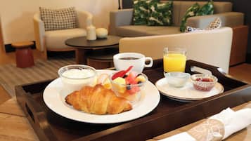 Bandeja de café da manhã com croissant, iogurte, frutas, café e suco em quarto de hotel, com sofá e uma mesa pequena.