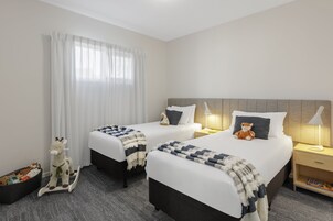 Quarto de hotel com duas camas, uma mesinha de cabeceira com abajur, uma pequena mesa com livros, um cavalinho de balanço e uma janela com cortinas brancas.