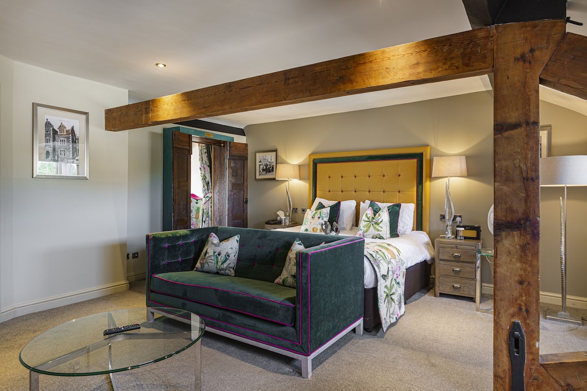 Quarto com sofá verde, cama com cabeceira de madeira, mesa de cabeceira e uma luminária.