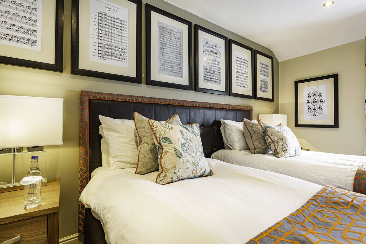 Quarto de hotel com duas camas, uma mesa de cabeceira com abajur, partitura emoldurada na parede e um quadro com pessoas.