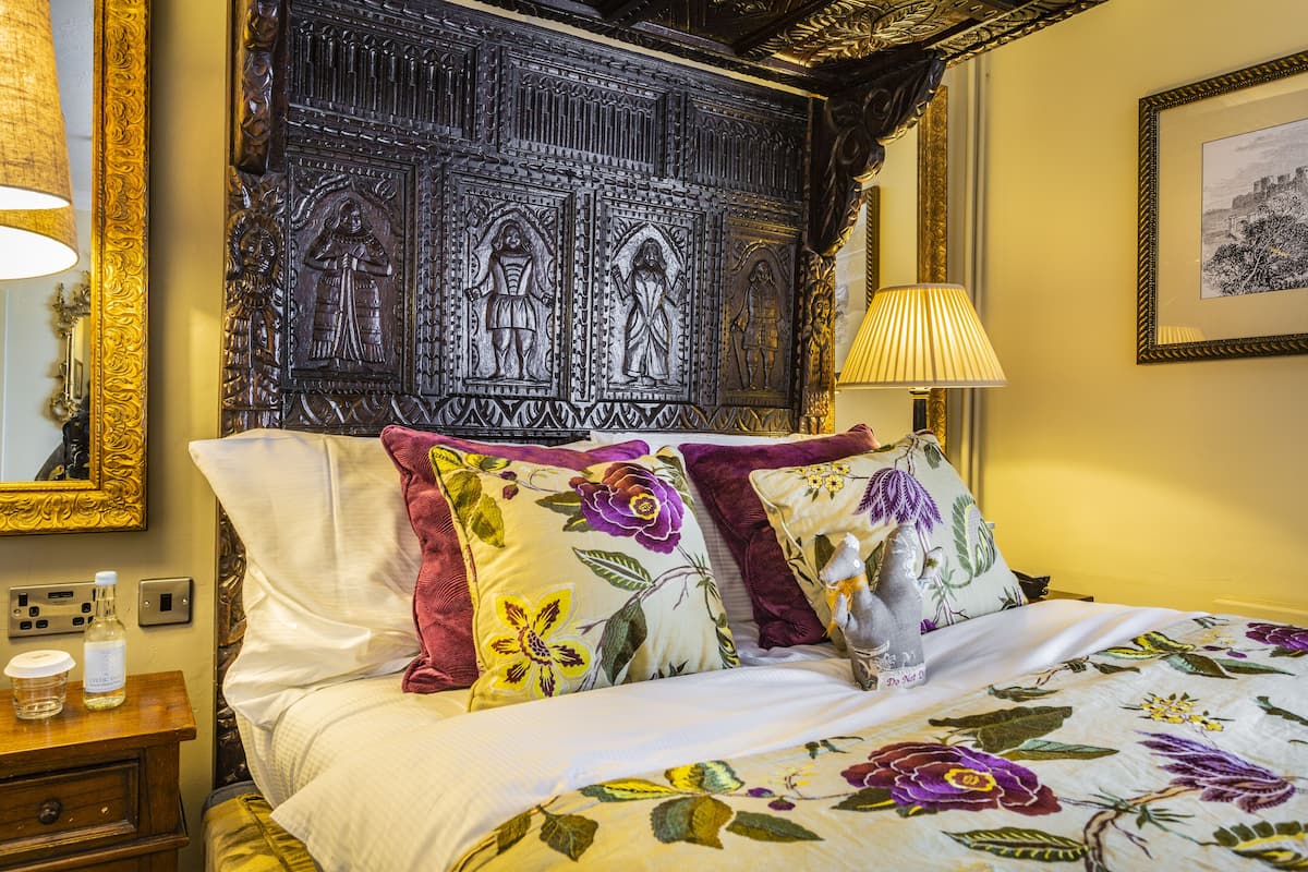 Cama com roupa de cama floral, cabeceira de madeira com entalhes, luminária de cabeceira e mesa de cabeceira com garrafa e copos.