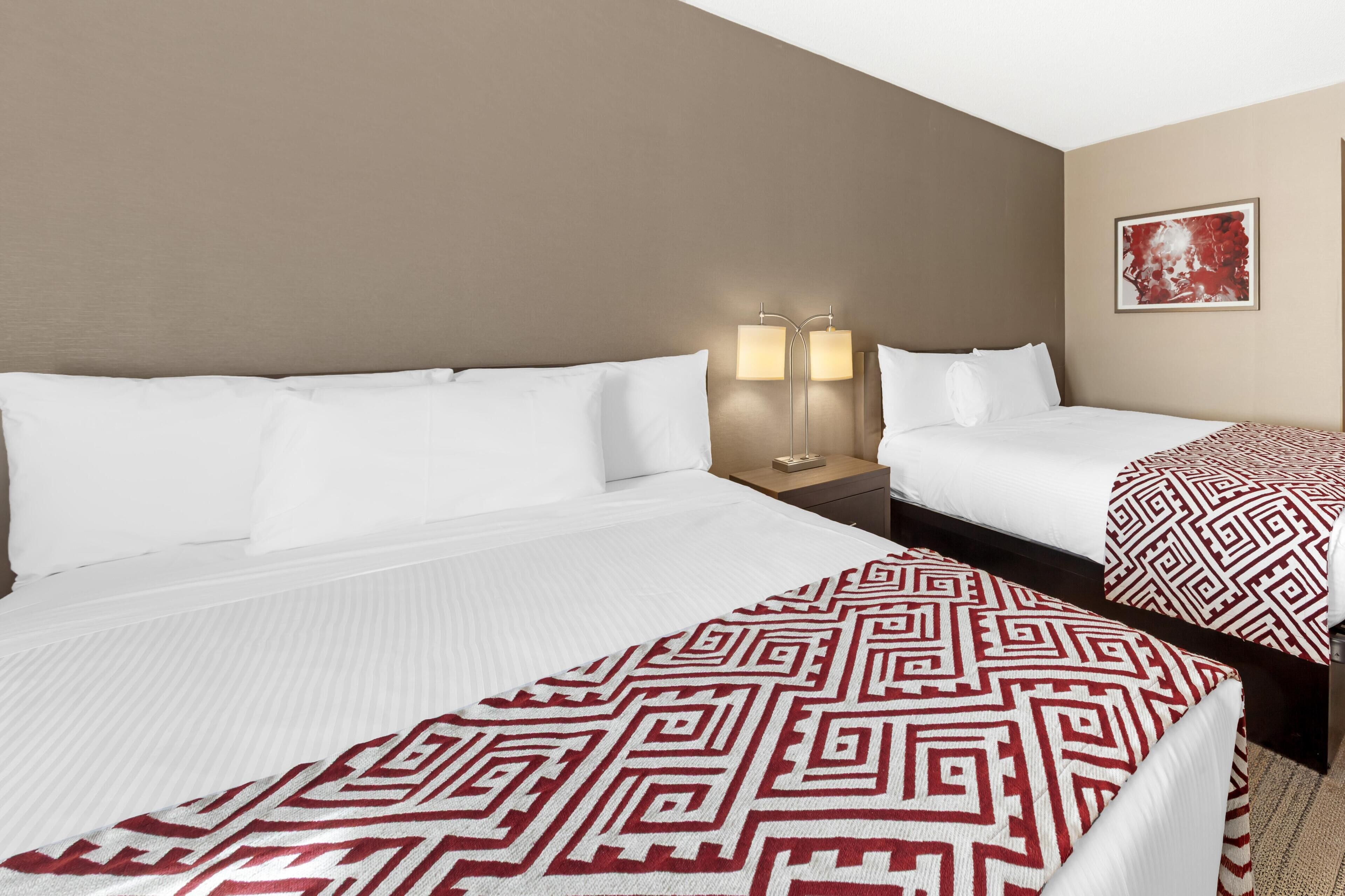 Quarto de hotel com duas camas, uma mesa de cabeceira com luminárias, um cobertor estampado e um quadro na parede.