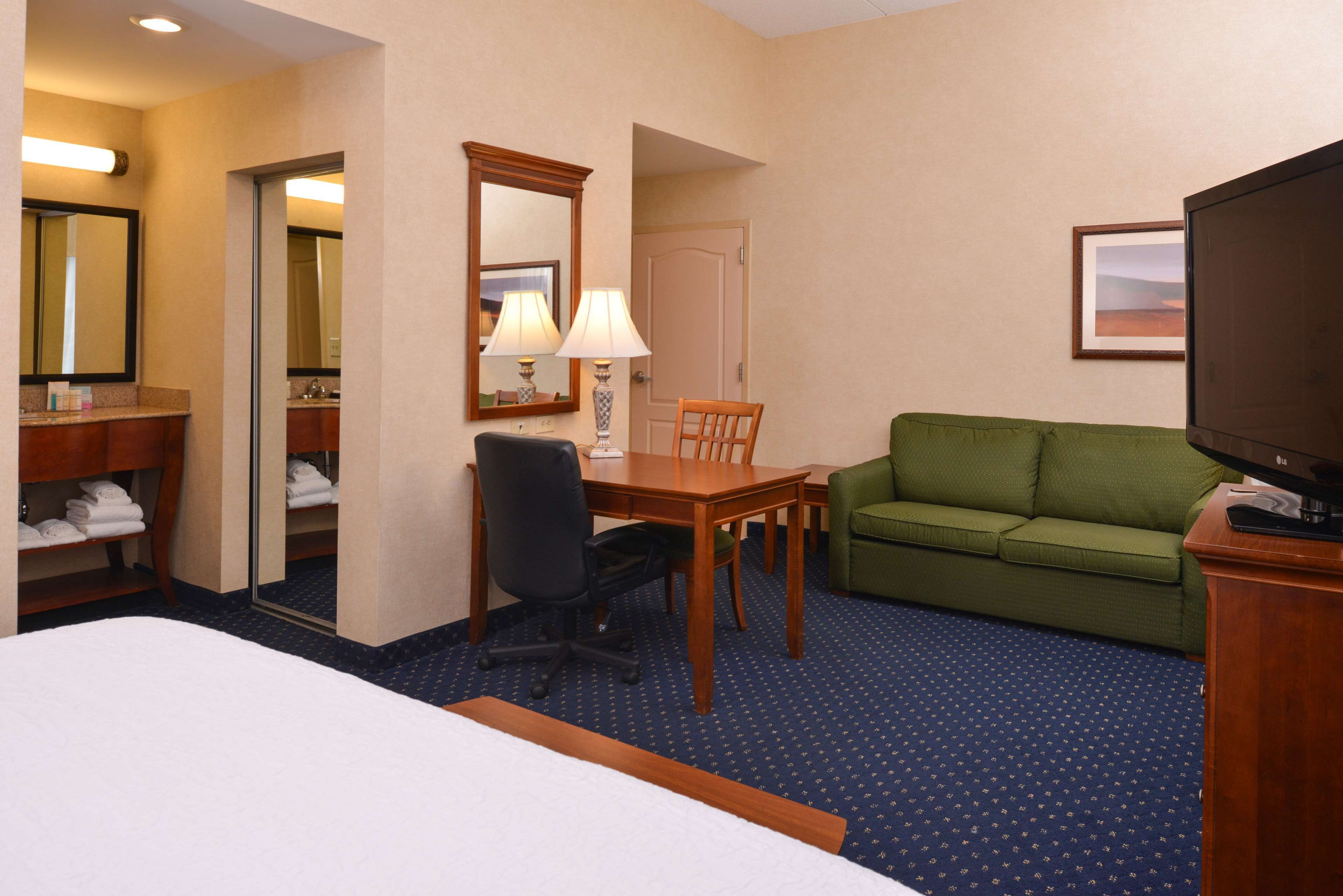 Quarto de hotel com uma cama, uma escrivaninha com cadeira, um sofá verde, uma televisão e um banheiro com espelho e pia.