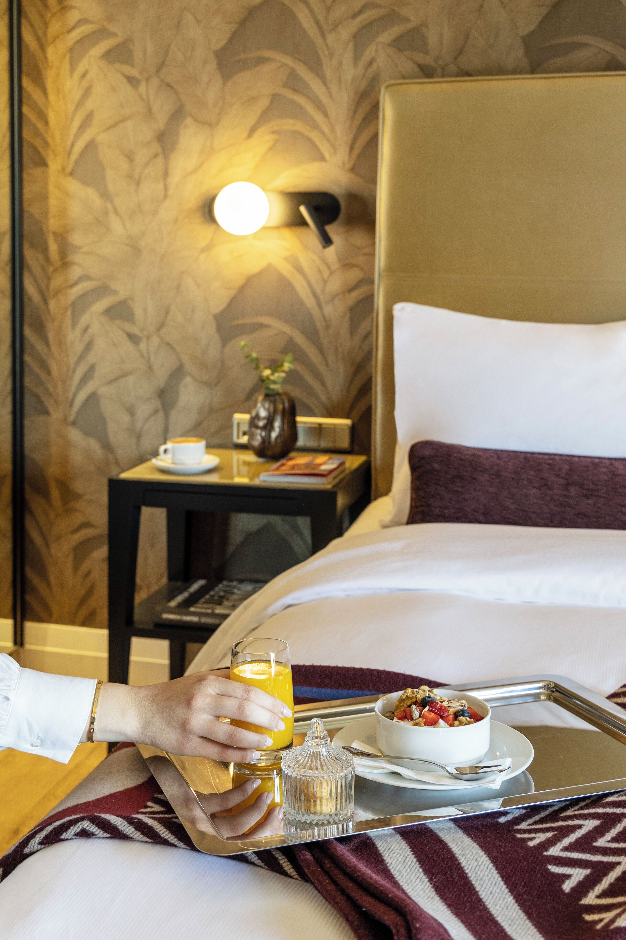 Quarto de hotel com uma cama, uma mesa de cabeceira com uma xícara e um vaso, e uma bandeja com tigela de cereal e um copo de suco de laranja.