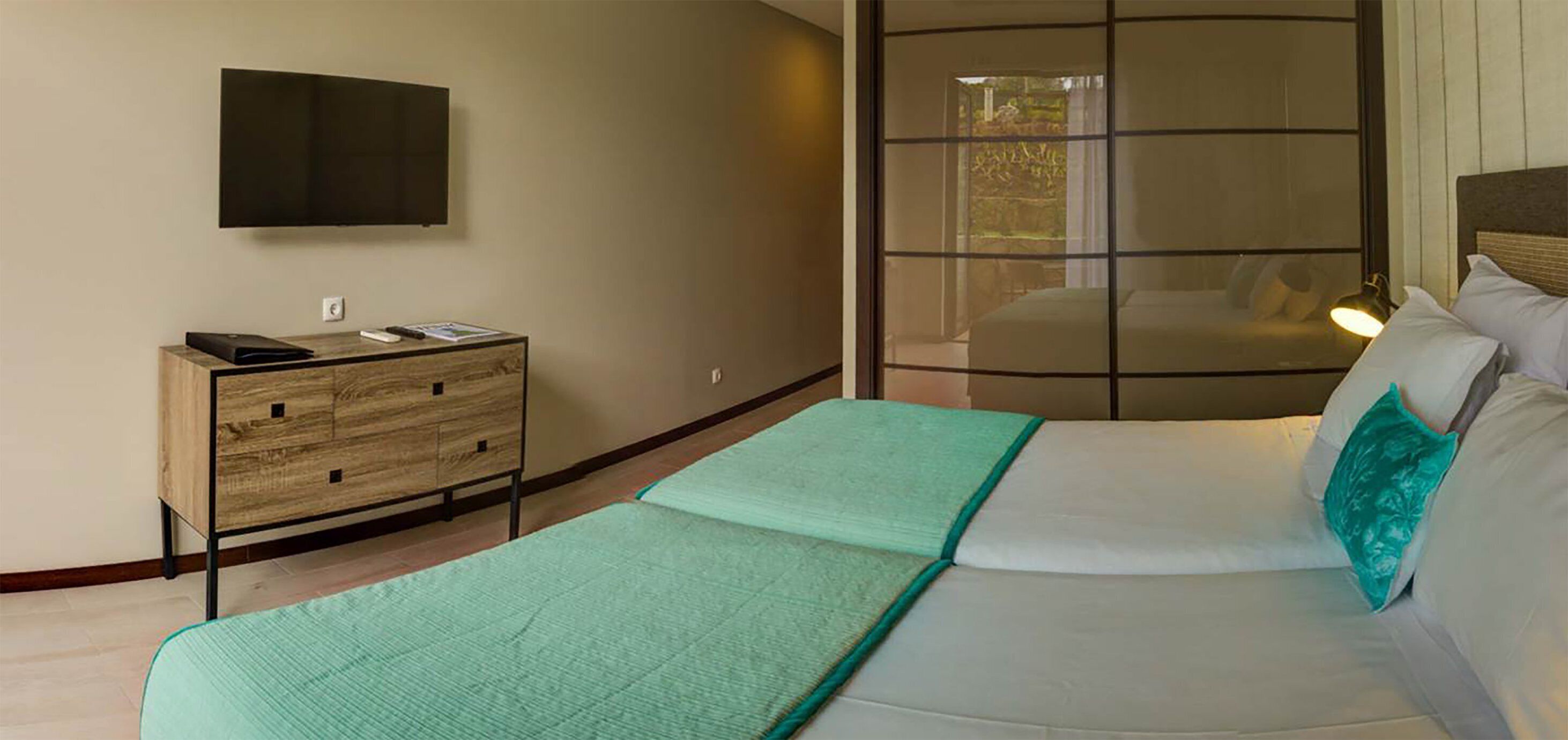 Quarto de hotel com cama, televisão fixada na parede, cômoda de madeira e uma janela grande.