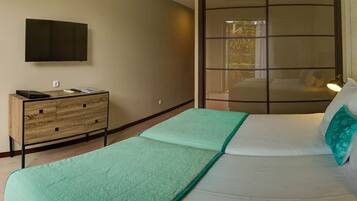 Quarto de hotel com cama, televisão fixada na parede, cômoda de madeira e uma janela grande.