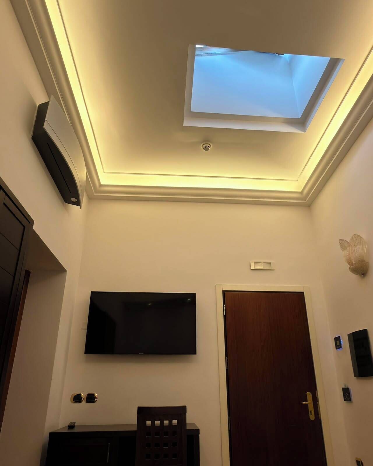 Quarto com claraboia, televisão de tela plana, porta de madeira e uma pequena mesa com cadeira.