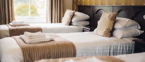 Quarto de hotel com duas camas, cada uma com almofada e toalha dobrada. O quarto possui uma janela com cortinas, uma cabeceira de madeira e uma parede com padrão de folhagem.