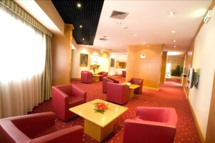 Um saguão de hotel com assentos vermelhos, uma mesa central com arranjo de flores e piso coberto por carpete vermelho.