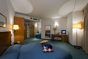 Quarto de hotel com cama, escrivaninha, cadeira e uma pequena mesa com uma planta.