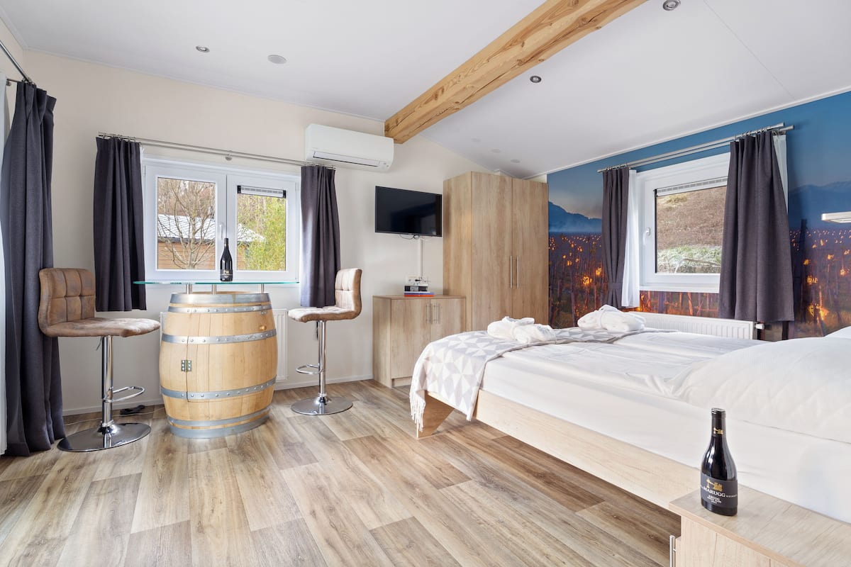Quarto de hotel moderno com teto de madeira, uma cama grande, televisão, minibar e um banquinho.