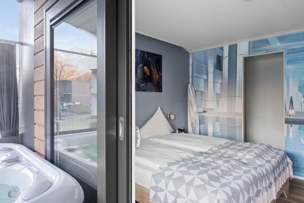 Quarto de hotel moderno com uma grande porta de vidro deslizante que dá para uma varanda, uma cama com manta de padrão geométrico, uma mesinha de cabeceira e um quadro pendurado na parede.